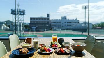 Uma mesa com diversos pratos, um copo de suco de laranja e uma vista para um campo esportivo e edifícios ao fundo.