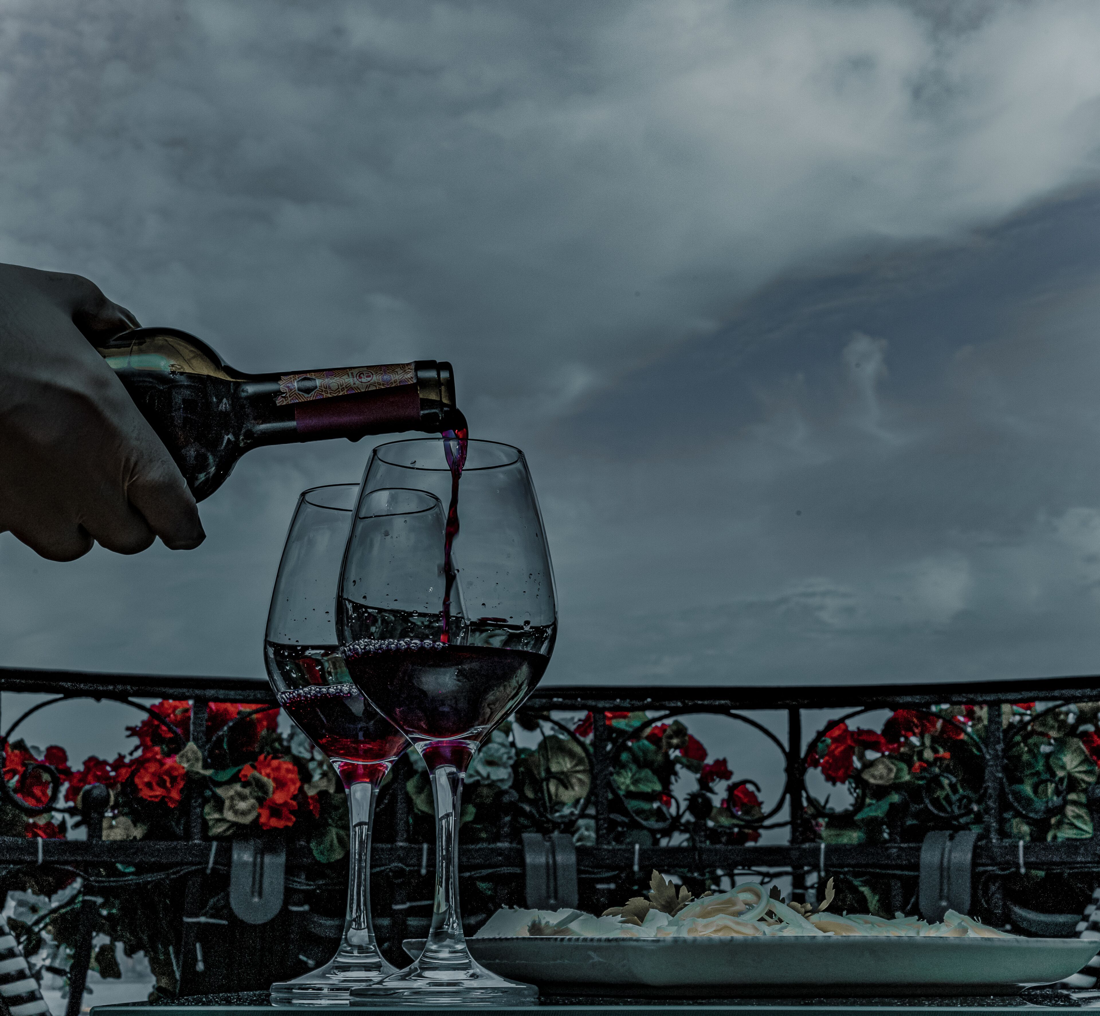 Uma mão servindo vinho tinto em um copo numa varanda com guarda-corpo e flores.