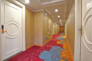Corredor de hotel com carpete vermelho estampado com folhas, portas brancas com puxadores circulares e iluminação embutida no teto.