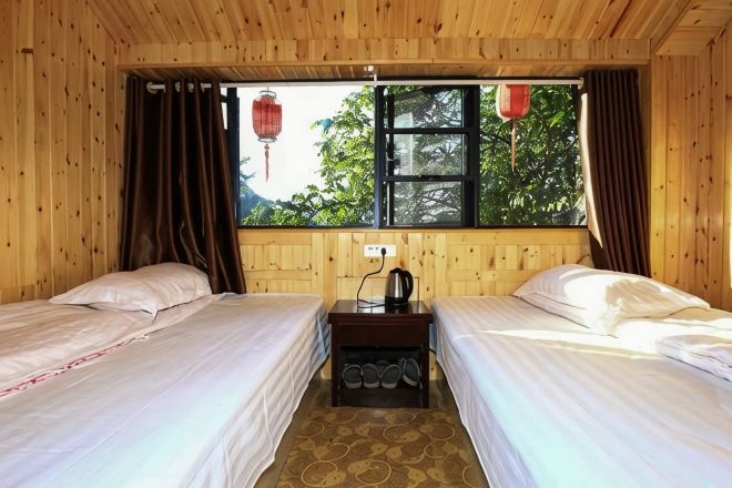 Quarto com duas camas, uma janela com cortinas, mesa de cabeceira com chaleira e tapete estampado.