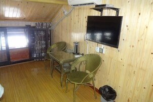Um quarto com painéis de madeira, mesa de vidro, cadeiras de vime, televisão na parede e uma janela com cortinas.