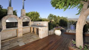 Cozinha externa com duas fornos de tijolo e uma pia, cercada por um deck de madeira e vegetação.