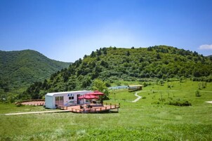 Uma cabana pequena e térrea com varanda e área de estar externa, rodeada por colinas verdejantes e céu claro.