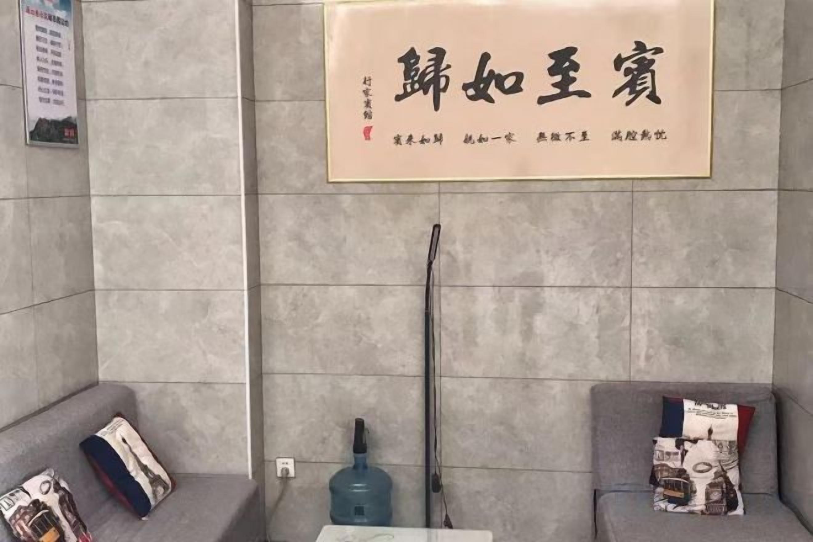 Um quarto com parede revestida de azulejos, um objeto longo preto no chão, dois sofás cinzas com almofadas e um cartaz com caracteres chineses.