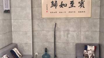 Um quarto com parede revestida de azulejos, um objeto longo preto no chão, dois sofás cinzas com almofadas e um cartaz com caracteres chineses.
