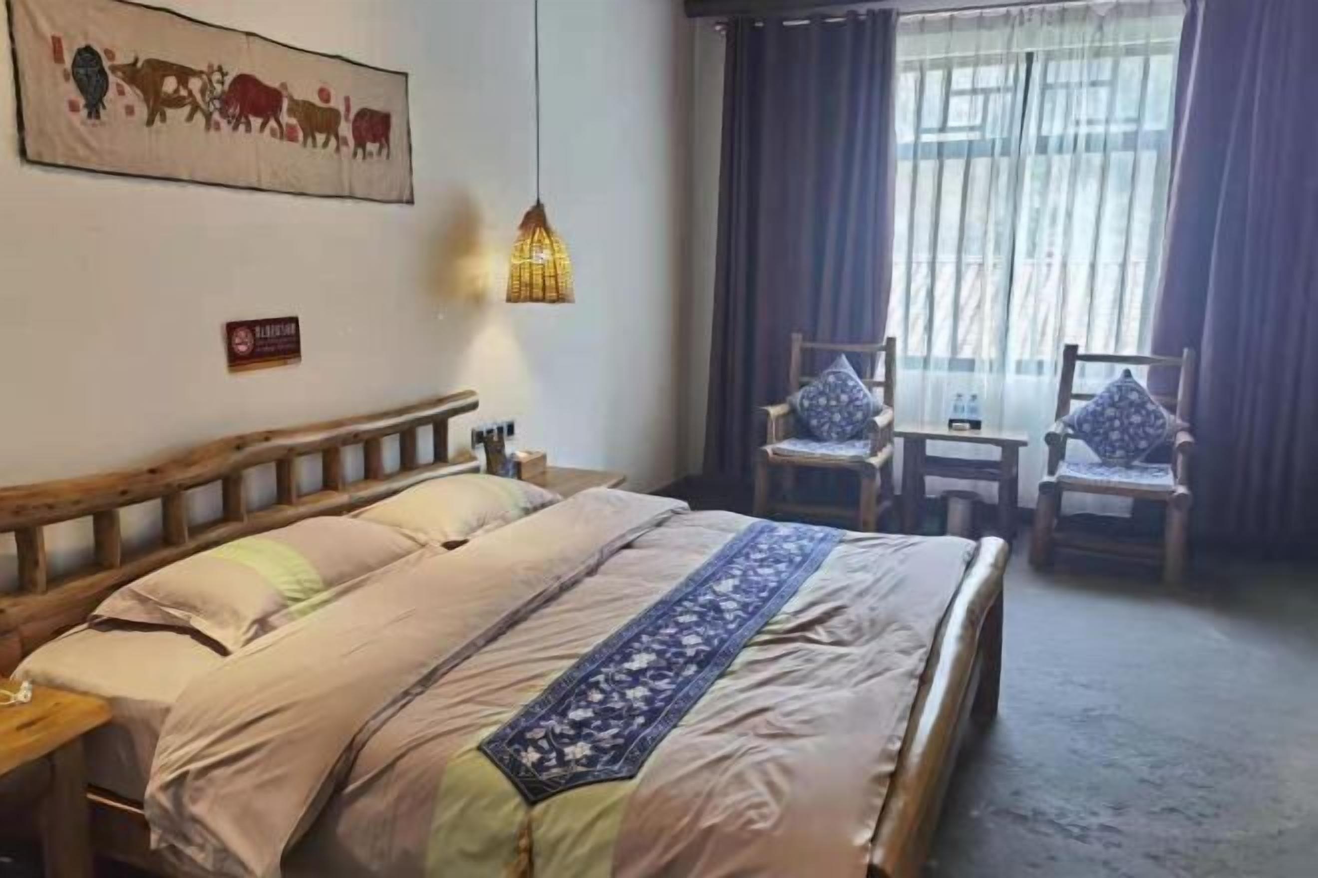 Uma cama com cabeceira, uma mesa de cabeceira, um abajur fixado na parede, duas cadeiras, uma janela com cortinas e um objeto decorativo na parede.