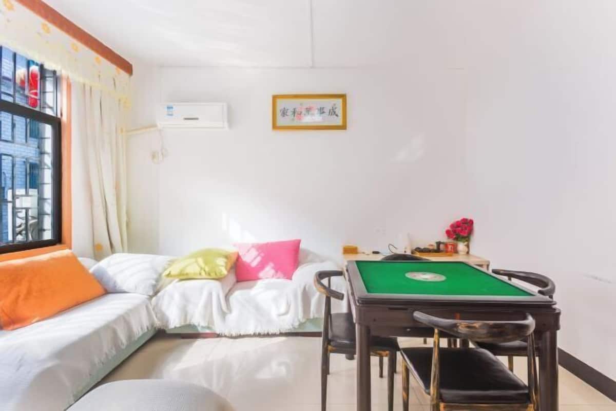 Um quarto com mesa de bilhar verde, sofá com almofadas laranja e rosa, e janela com cortinas.