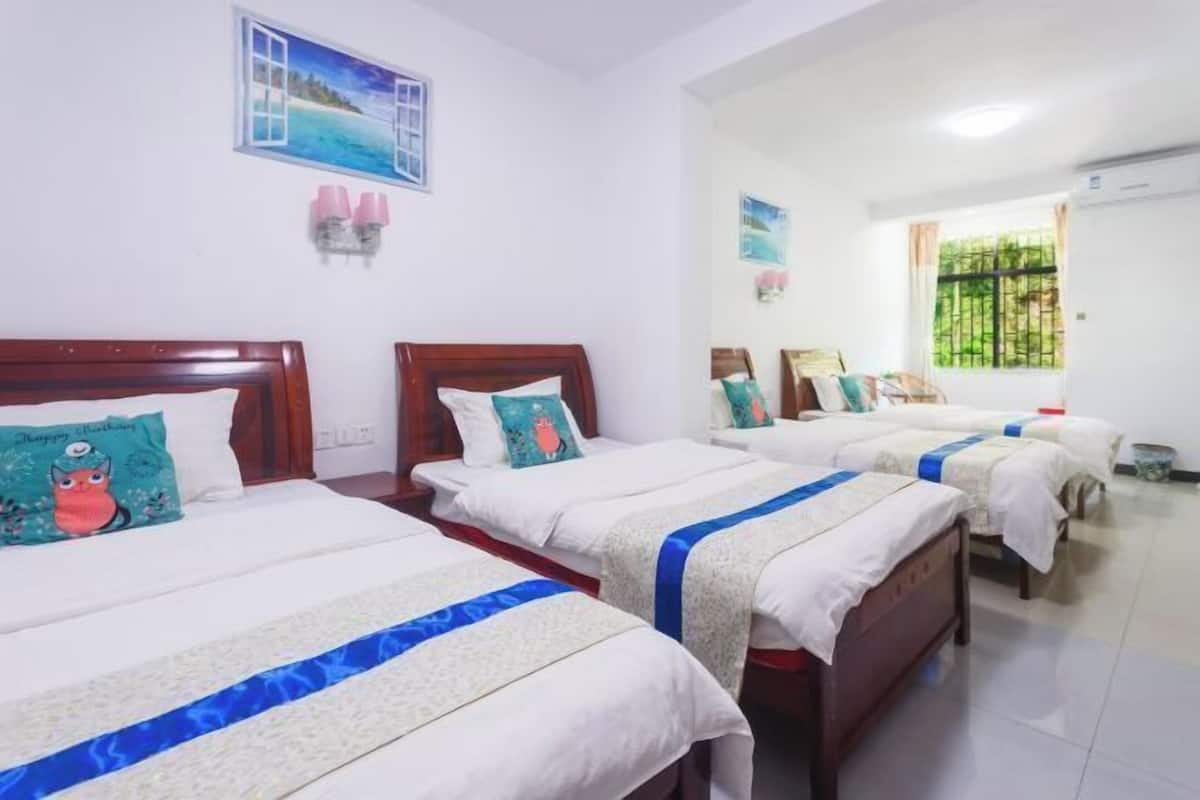 Quarto de hotel com quatro camas, cada uma com cabeceira de madeira e roupa de cama branca. Há luminárias fixadas na parede e uma janela com vista para o verde.