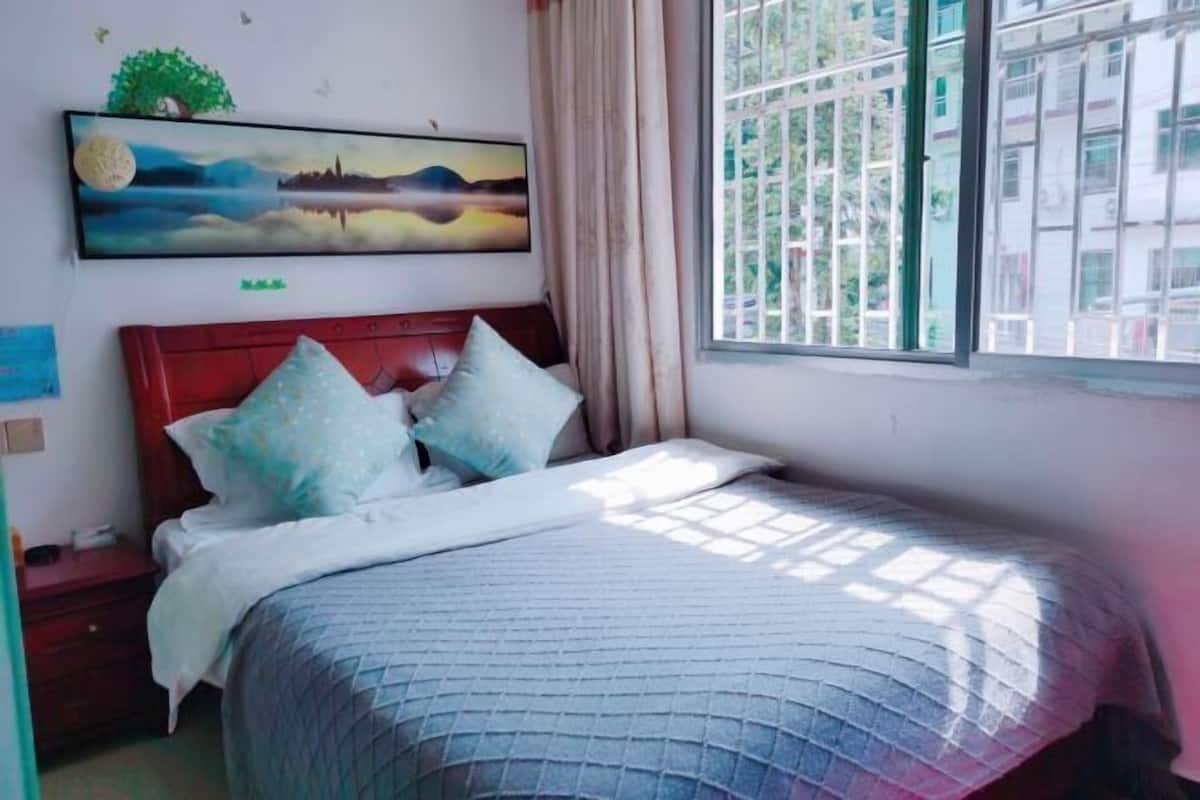 Quarto com cama, mesa de cabeceira, janela com vista para edifícios e um quadro na parede.