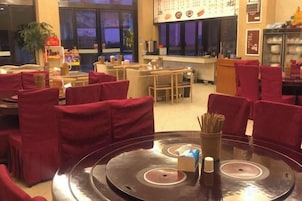 Um restaurante com mesas redondas, cadeiras estofadas vermelhas e uma área de buffet ao fundo.