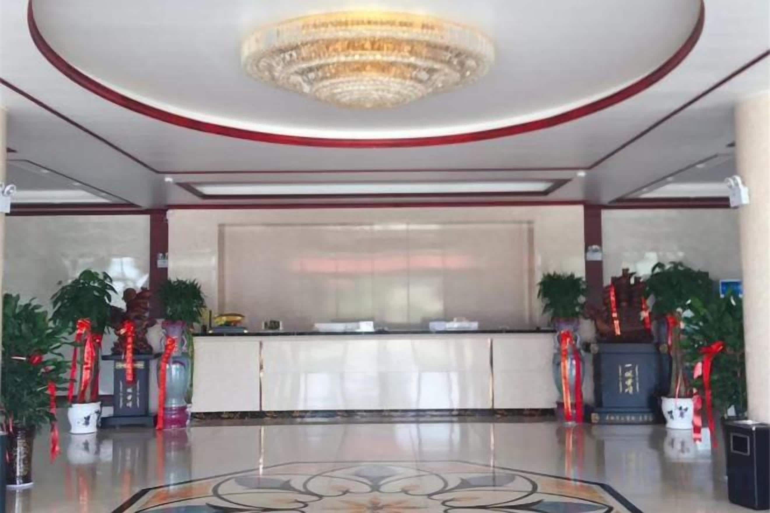 Um hall de hotel com balcão de recepção, lustre e plantas em vasos.
