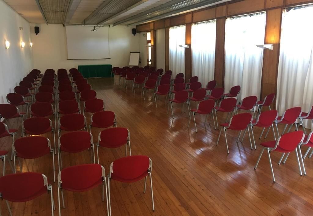 Uma sala de conferências com fileiras de cadeiras vermelhas, uma tela de projeção e um quadro branco.