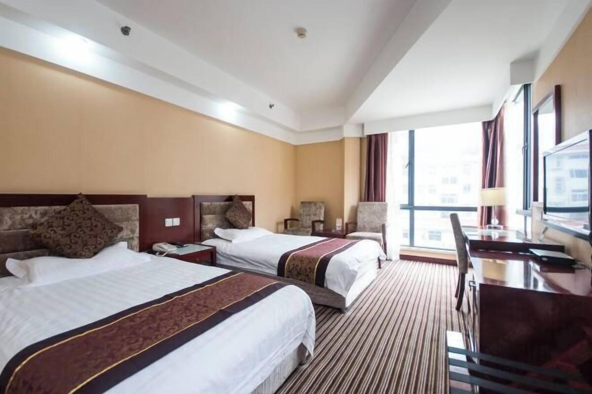 Quarto de hotel com duas camas, uma escrivaninha, uma cadeira e uma janela grande.