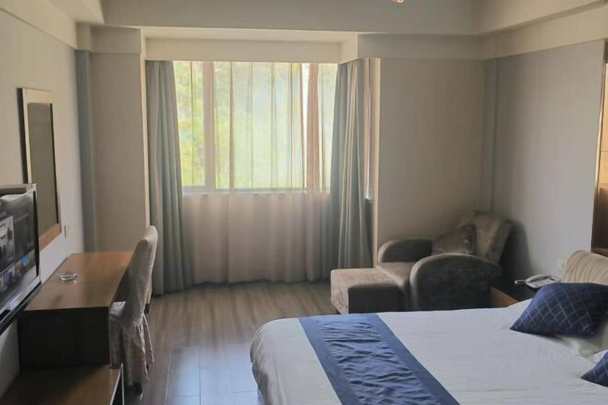 Quarto de hotel com cama, escrivaninha, cadeira, televisão e janela com cortinas.