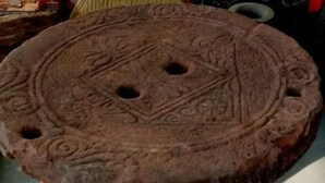 Um artefato de pedra com esculturas detalhadas e quatro furos, montado em um suporte de madeira.