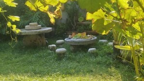 Um jardim com mesas de pedra e uma fonte, rodeado por vegetação e plantas.