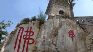Uma grande rocha com caracteres chineses entalhados, um edifício tradicional com telhado de telhas e um céu azul claro ao fundo.