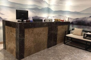 Uma mesa de recepção com monitor de computador, telefone e uma pequena mesa com um vaso de flores. Há um banco com almofada em primeiro plano.