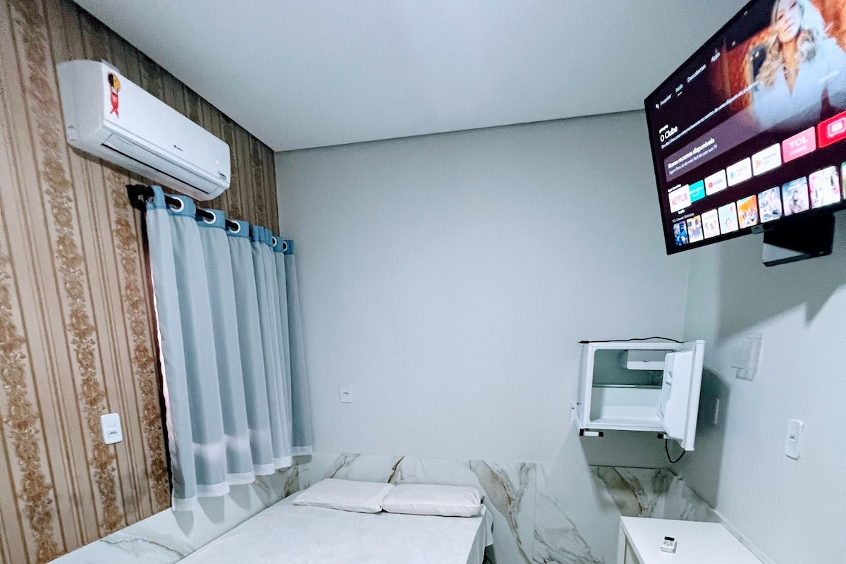 Quarto de hotel com cama, televisão na parede, ar condicionado e uma mesinha.