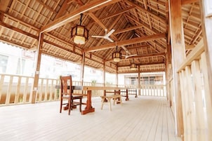 Um pavilhão ao ar livre com vigas de madeira, ventiladores de teto e mesas e cadeiras.