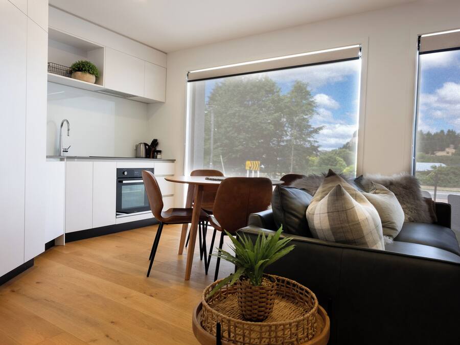 Cozinha moderna com armários brancos, área de refeições com mesa e cadeiras de madeira, e um sofá preto com almofadas decorativas.