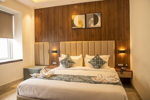 Quarto de hotel com uma cama grande, duas luminárias fixadas na parede, uma cabeceira de madeira e uma janela com persiana.