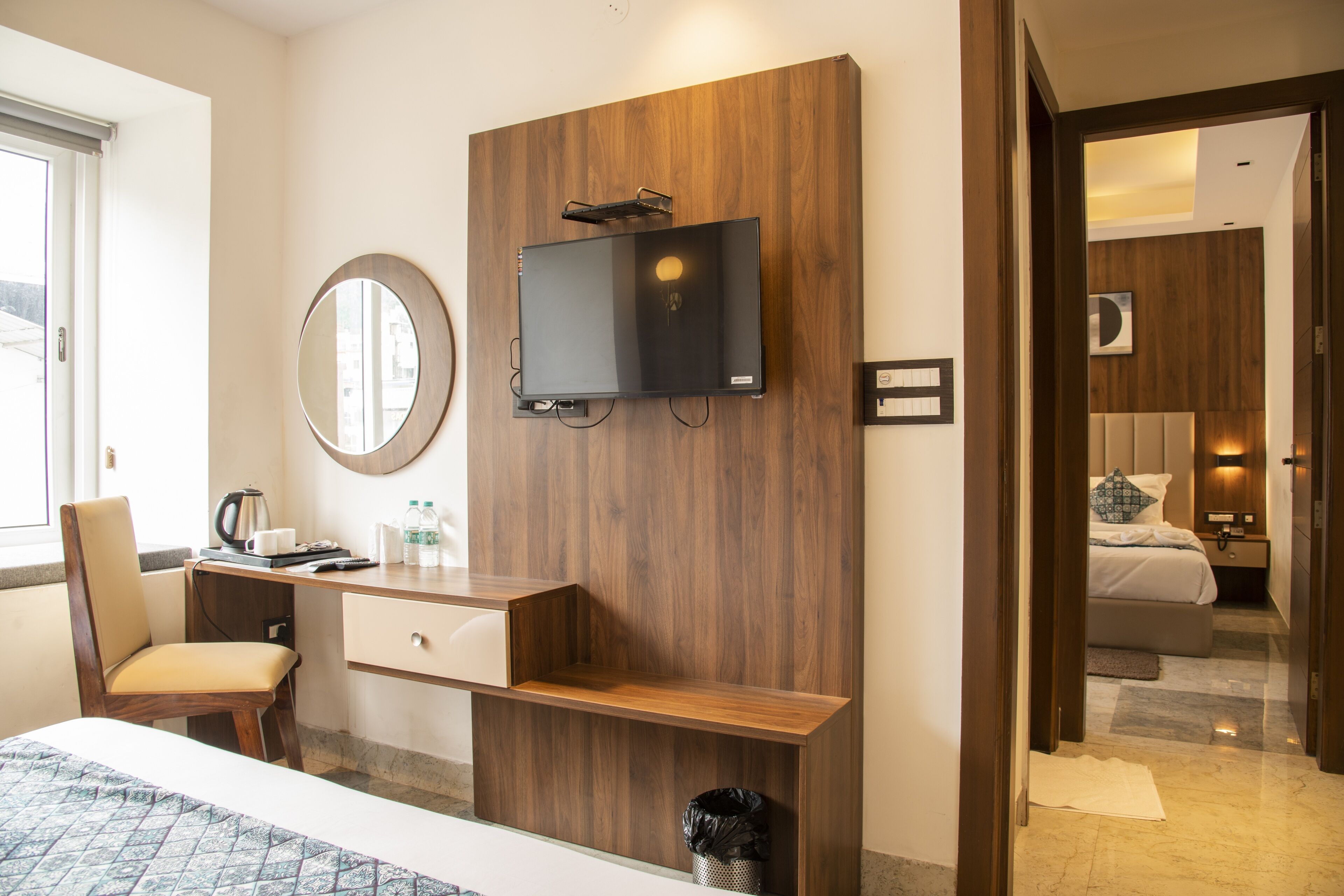 Quarto de hotel com uma cama, uma escrivaninha com cadeira, uma televisão fixada em um painel de madeira, um espelho redondo e uma pequena lixeira.