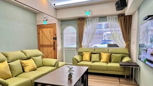 Um quarto com dois sofás verdes, uma porta de madeira, uma janela com cortinas e uma pequena mesa com uma planta.