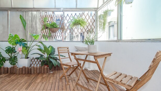 Um deck de madeira com mesa e cadeiras, rodeado por plantas em vasos e um painel de madeira treliçada.