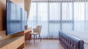 Quarto de hotel moderno com uma televisão de tela plana grande presa na parede, uma cama confortável, um espaço de estar com uma poltrona e janelas amplas com cortinas leves.