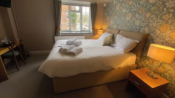 Uma cama bem arrumada com toalhas, uma mesinha de cabeceira com abajur, uma janela com cortinas e um padrão de papel de parede floral.