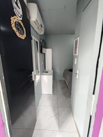 Um corredor estreito com uma pia branca, um espelho e uma cama com cobertor cinza.