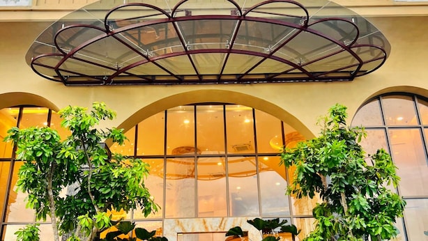 A entrada do hotel MAIVY, com uma cobertura de vidro, degraus de mármore e plantas em vasos.