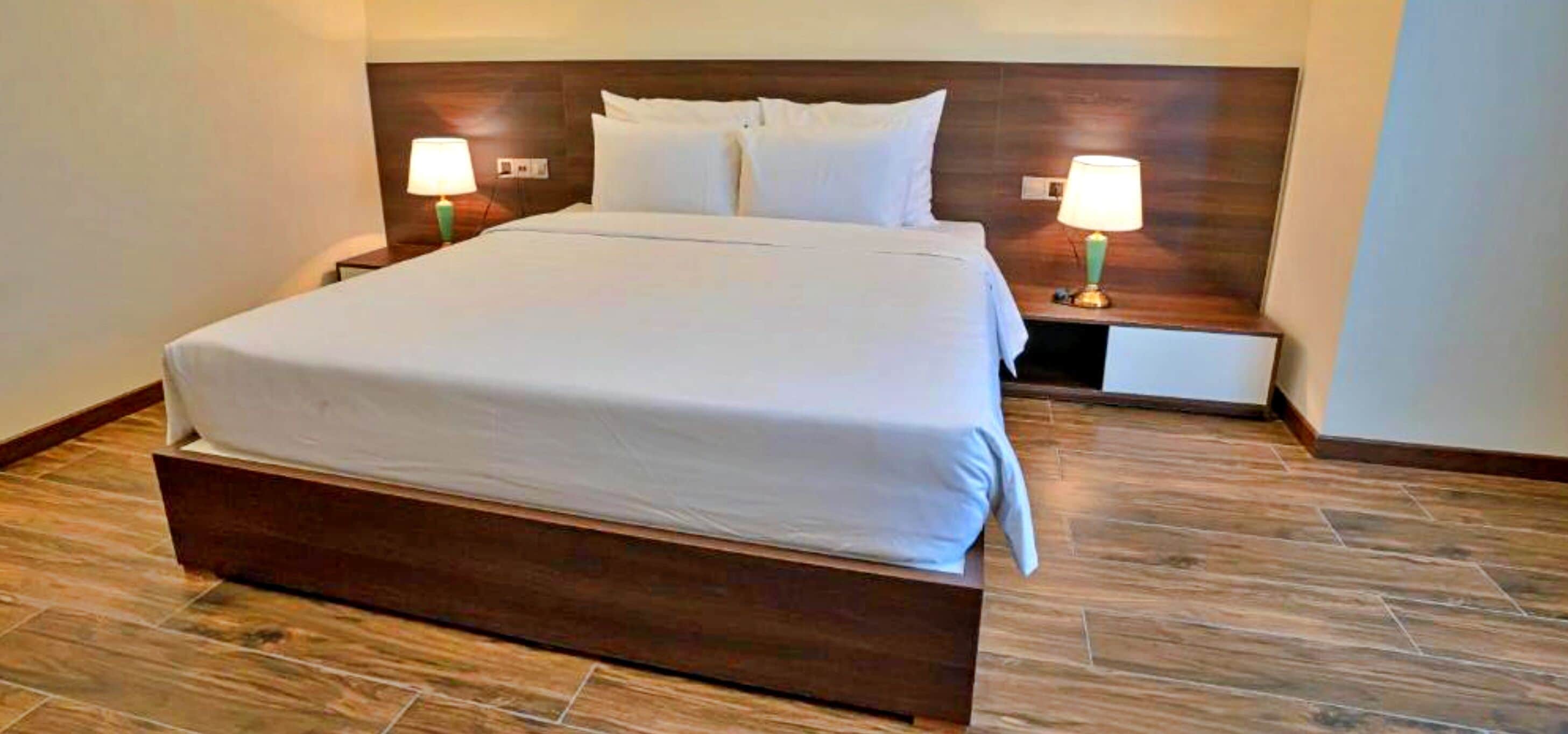 Uma cama grande com cabeceira de madeira, duas mesas de cabeceira com luminárias e piso de madeira.