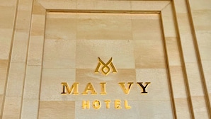 Placa de hotel com o nome 'MAI VY HOTEL' em letras douradas sobre uma parede de mármore.