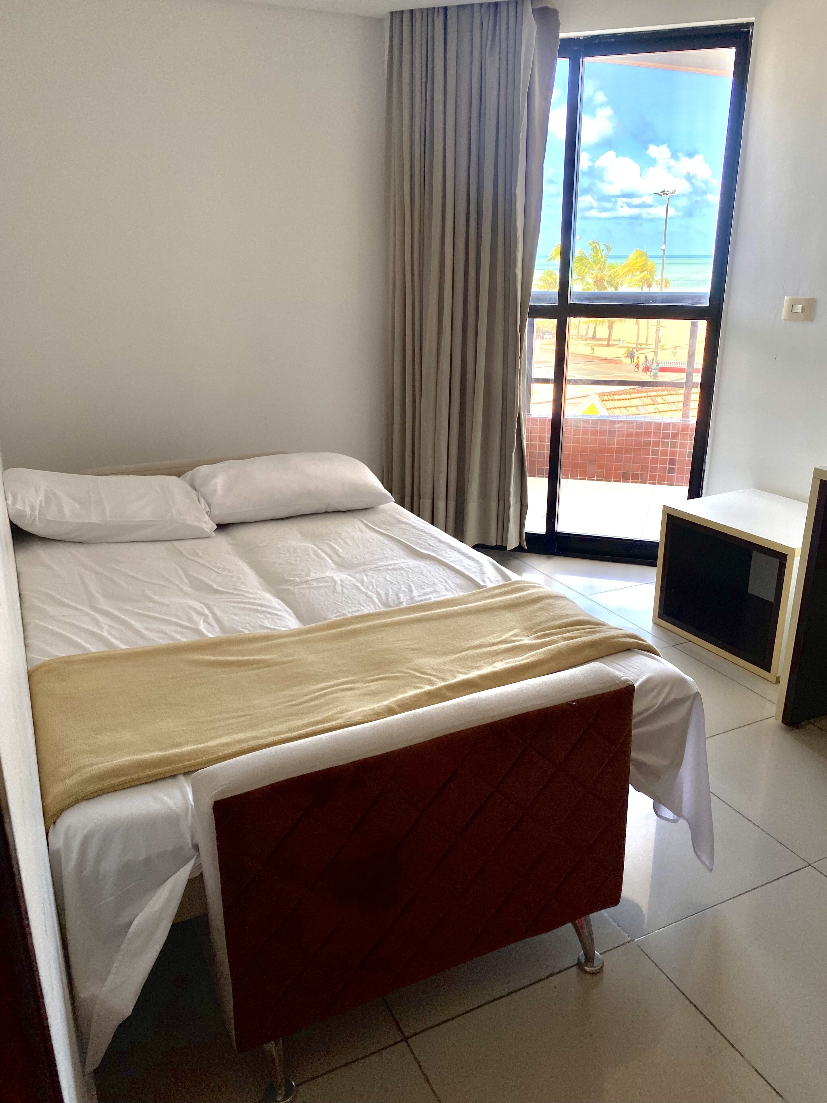 Quarto de hotel com cama, janela com vista para a praia e uma mesinha.