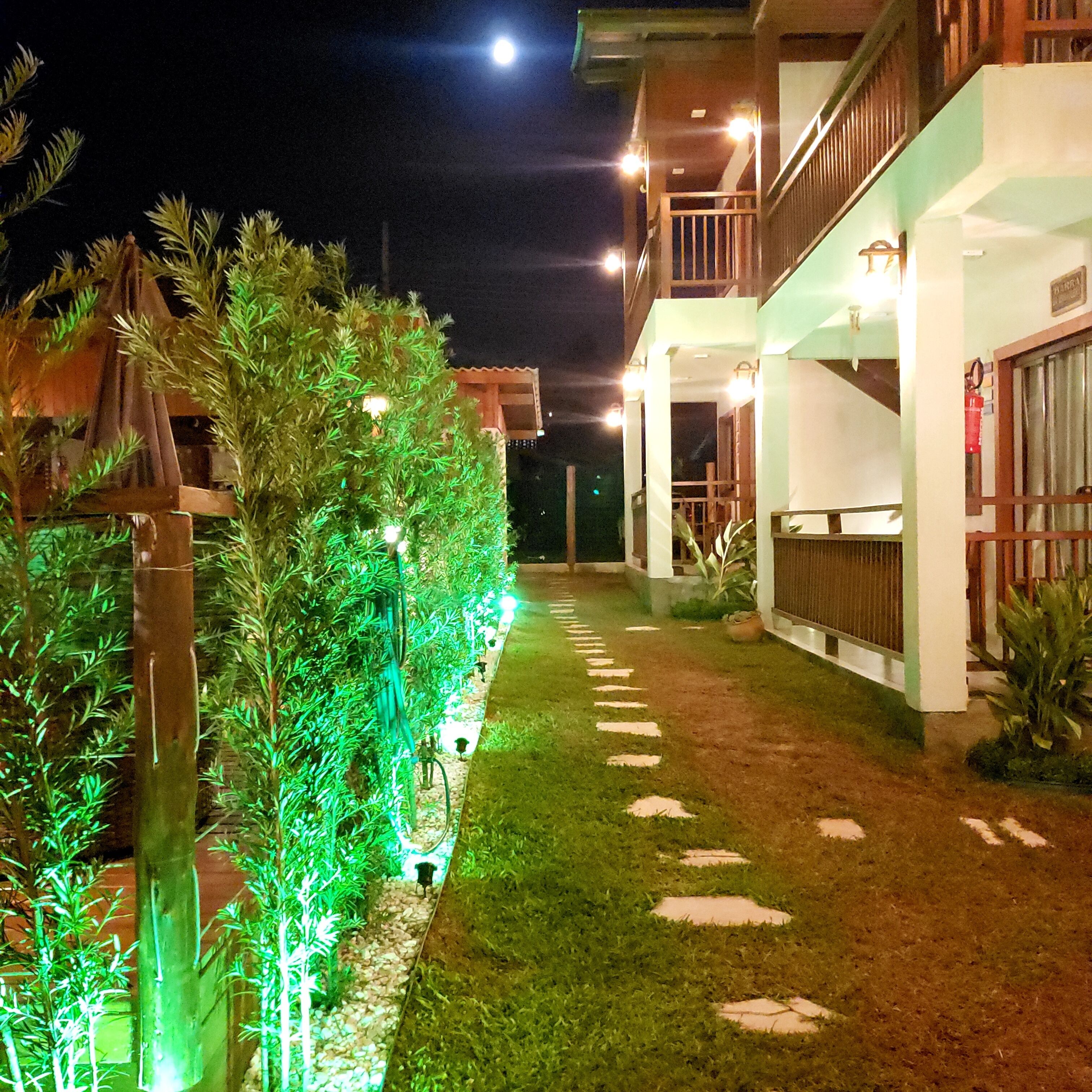 Caminho bem iluminado que leva a um edifício com varanda, cercado por vegetação e degraus de pedra.