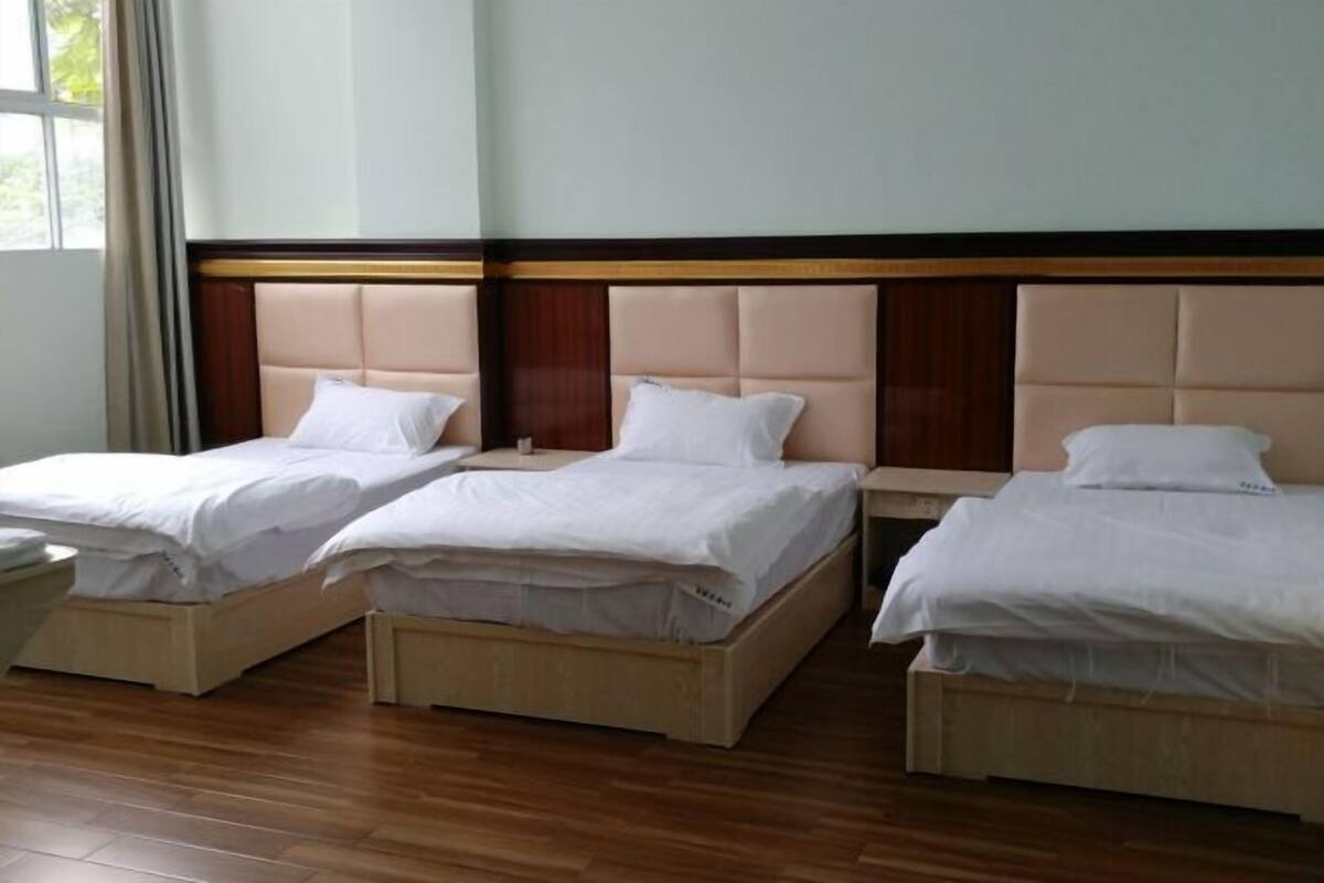 Quarto de hotel com três camas de solteiro, cabeceiras de madeira e uma janela grande.