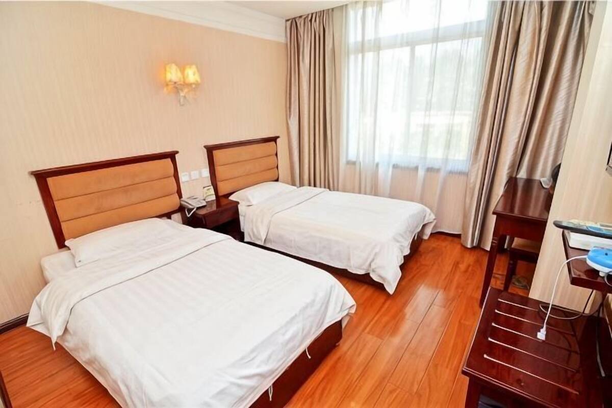 Quarto de hotel com duas camas, piso de madeira, uma escrivaninha e janela com cortinas.