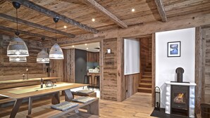 Um interior rústico com uma mesa de jantar de madeira, bancos, luminárias pendentes, uma lareira e uma escada que leva a outro cômodo.