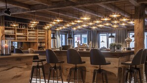 Uma área de bar com balcões de madeira, vários bancos, luminárias pendentes e prateleiras com garrafas e copos.