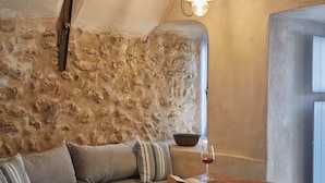 Um quarto aconchegante com parede de pedra, uma mesa de madeira com um livro e um copo, e um sofá com almofadas listradas.