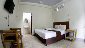 Quarto de hotel com cama, uma cadeira de madeira, uma mesinha, televisão fixada na parede e banheiro com pia e espelho.