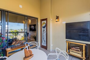 Quarto com televisão grande, mesa de madeira, cadeiras, porta de vidro que dá para o exterior e luminária fixada na parede.