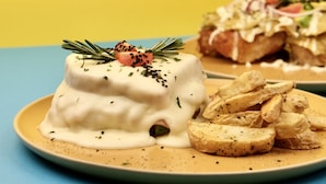 Um prato de sobremesa com um doce cremoso branco, decorado com ervas e um raminho de alecrim, acompanhado de batatas assadas.