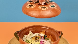 Uma mão erguendo a tampa de um pote de barro com desenho floral, revelando um prato com arroz, legumes e uma porção de molho em um prato.