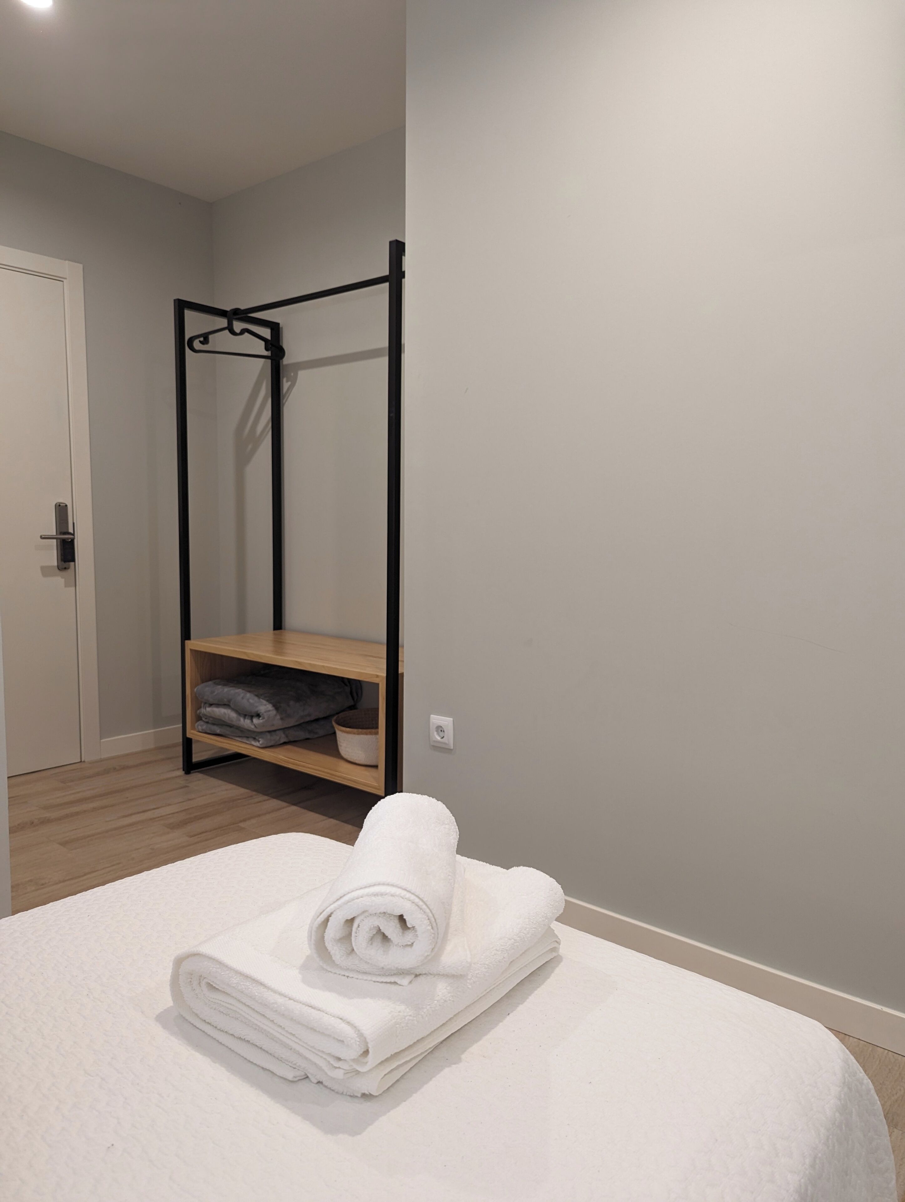 Cama bem arrumada com toalhas brancas, prateleira de madeira com toalhas dobradas e estrutura de cama de metal preto em um quarto moderno com paredes de cores claras e piso de madeira.