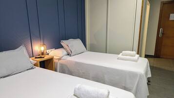 Quarto de hotel com duas camas de solteiro, mesa de cabeceira com abajur, guarda-roupa e uma porta.