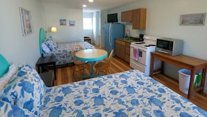 Quarto compacto com cama, uma mesinha, cozinha compacta e janela.