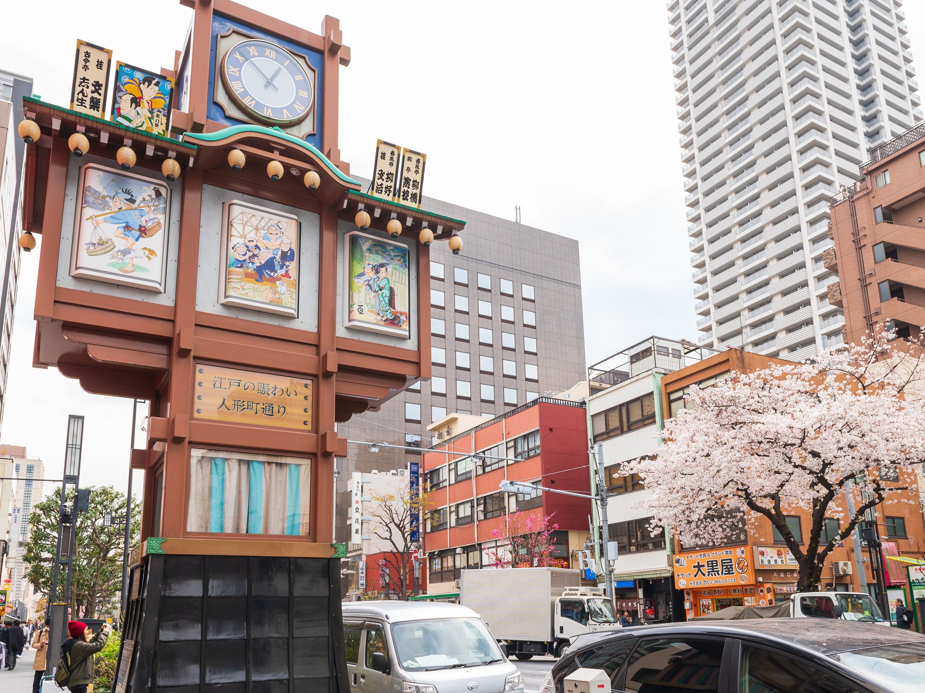 Um edifício tradicional japonês com várias placas e um relógio, rodeado por arranha-céus modernos e cerejeiras em flor.